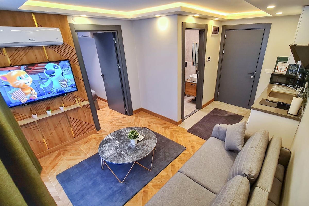 Uma sala de estar moderna com televisão de tela plana, uma mesa de centro redonda, um sofá cinza e uma cozinha visível através de uma porta aberta.