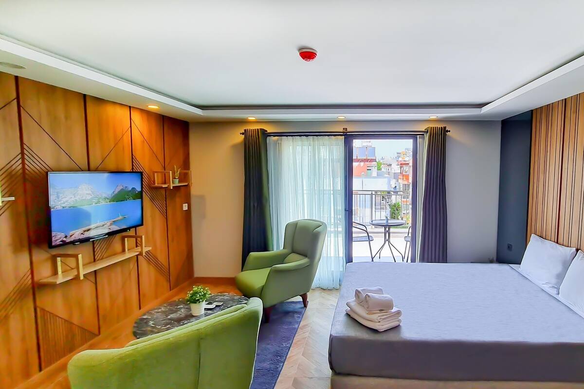 Quarto de hotel com cama, televisão na parede, cadeira e uma mesinha com uma planta.