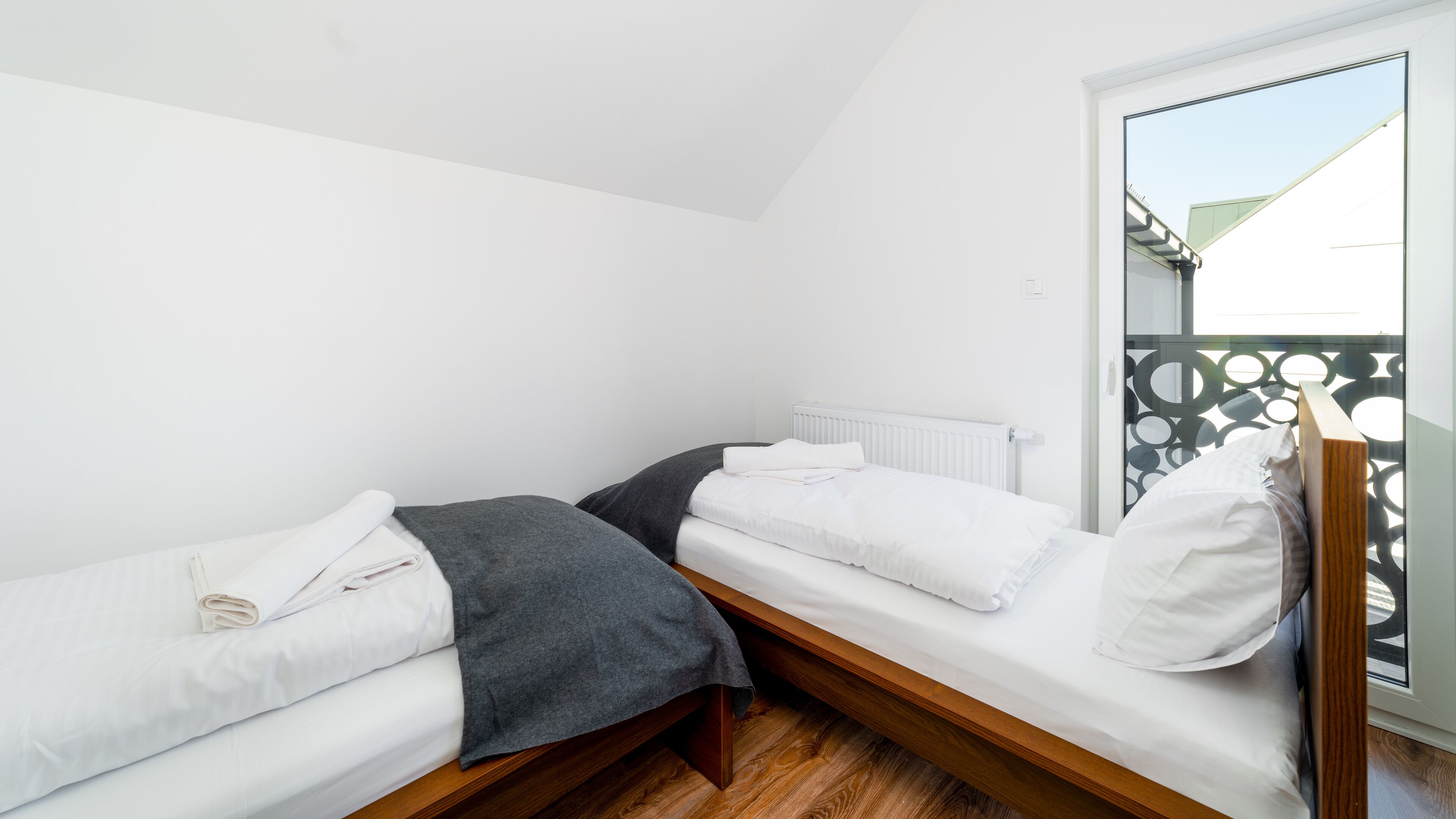 Quarto pequeno e moderno com duas camas individuais, um radiador e uma janela com vista para um edifício.