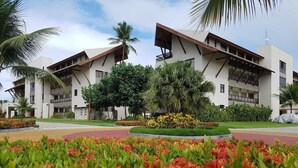 Hotel moderno com fachada simétrica, diversas varandas e paisagismo exuberante com plantas vermelhas e verdes.