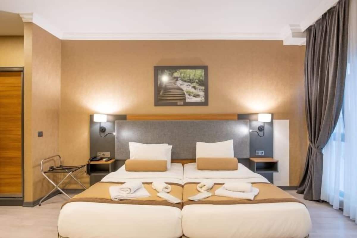 Quarto de hotel com uma cama de casal, duas mesas de cabeceira, cabeceira, um quadro e uma janela com cortinas.