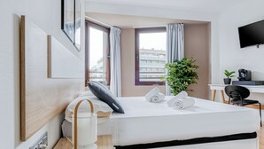 Quarto de hotel com cama, escrivaninha, televisão, janela com cortinas, uma planta e vista para edifícios.