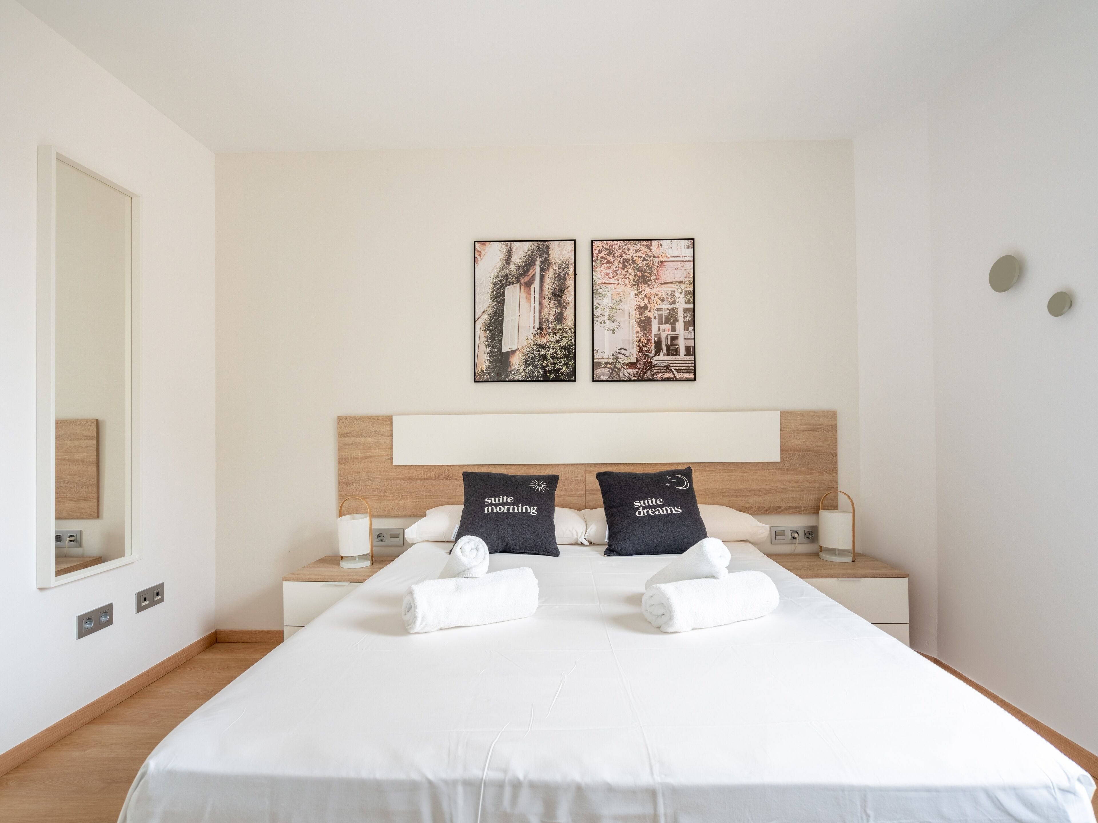 Quarto de hotel com uma cama grande, duas mesas de cabeceira, um criado-mudo, um espelho e duas obras de arte na parede.