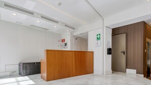 Uma área de recepção moderna com um balcão de madeira, parede branca com equipamentos de emergência e piso de mármore.