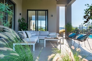 Uma área de estar externa moderna com sofá, mesa de centro e cadeiras, cercada por plantas e com vista para uma piscina.