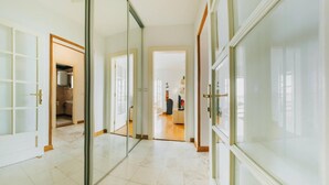 Um corredor amplo com uma porta de vidro que dá para um quarto, uma porta de madeira para outro quarto e vista para uma cozinha.