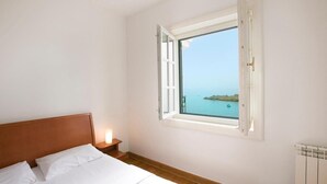 Quarto com cama, mesa de cabeceira, janela com persiana aberta e vista para um corpo d'água.