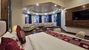 Quarto de hotel moderno com uma cama grande, sofá seccional, televisão na parede e vista da cidade pelas janelas.