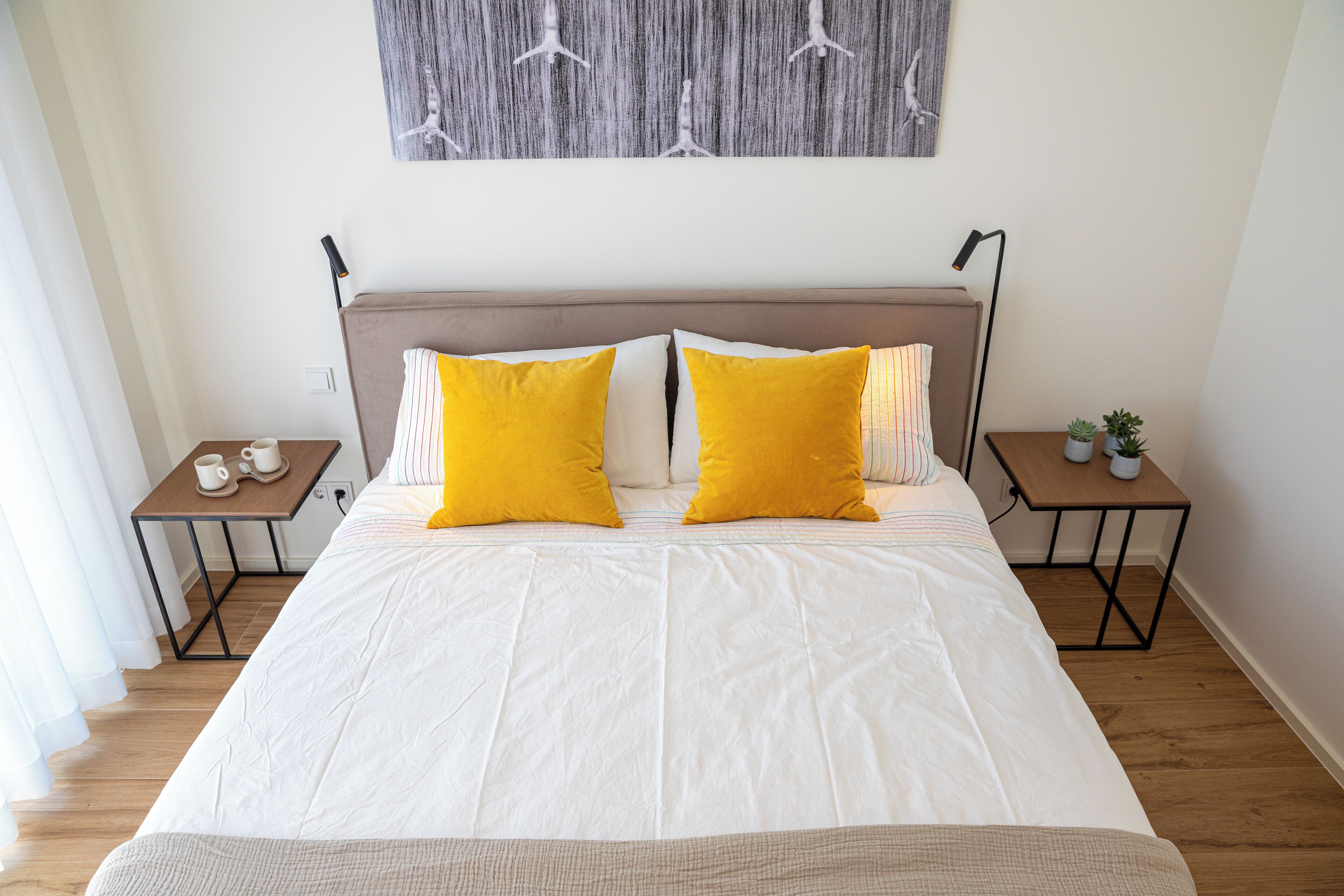 Cama bem arrumada com roupa de cama branca e amarela, duas mesas de cabeceira com abajures, e uma pequena planta em vaso sobre uma delas.