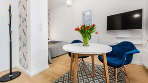 Uma sala de estar moderna com uma mesa redonda branca, uma cadeira azul, uma TV fixada na parede e um vaso com flores vermelhas sobre a mesa.