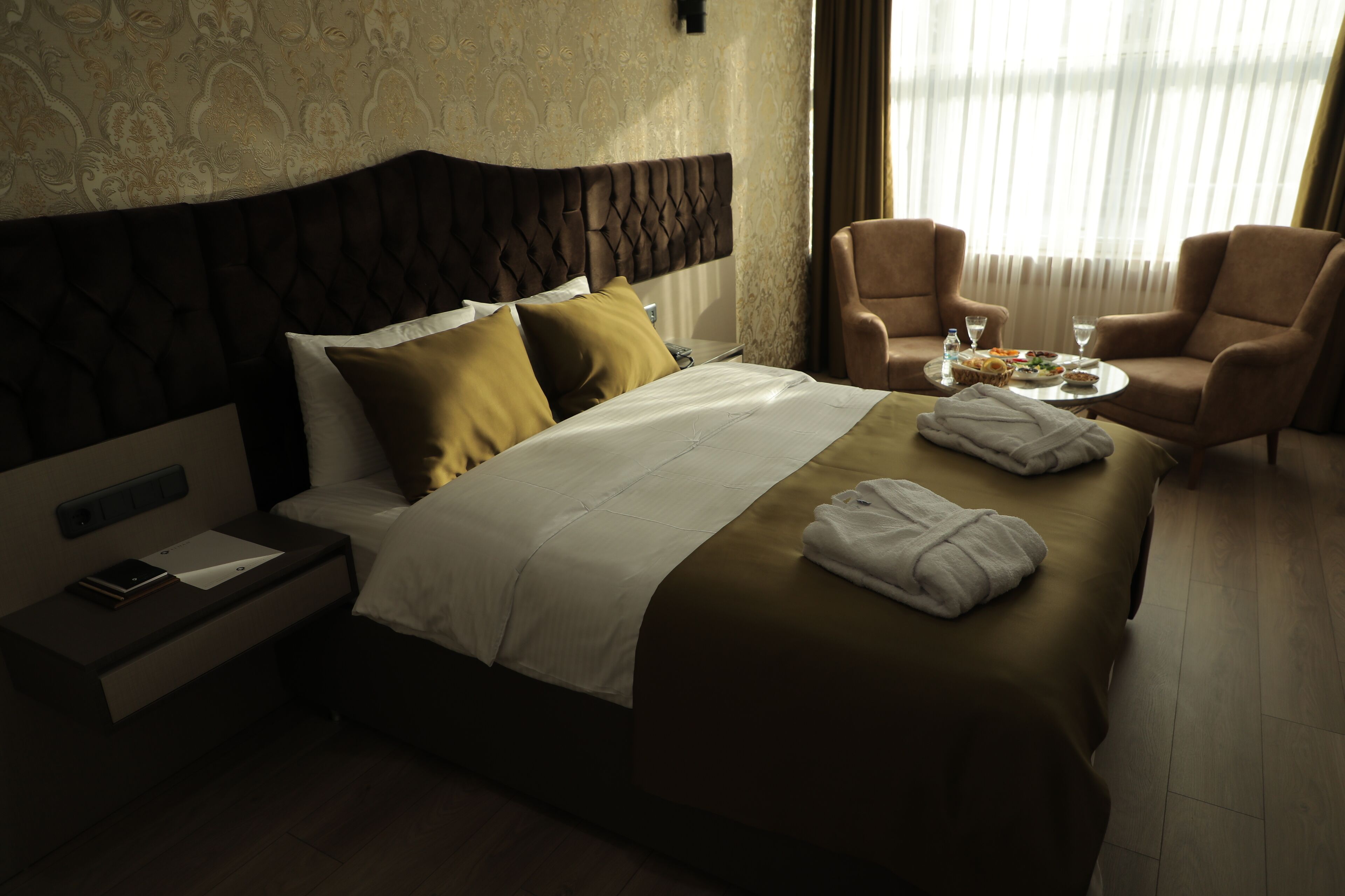 Quarto de hotel com uma cama grande, duas poltronas, uma mesinha de centro com um tabuleiro de comida e bebidas, e uma mesa de cabeceira com um telefone e papéis.