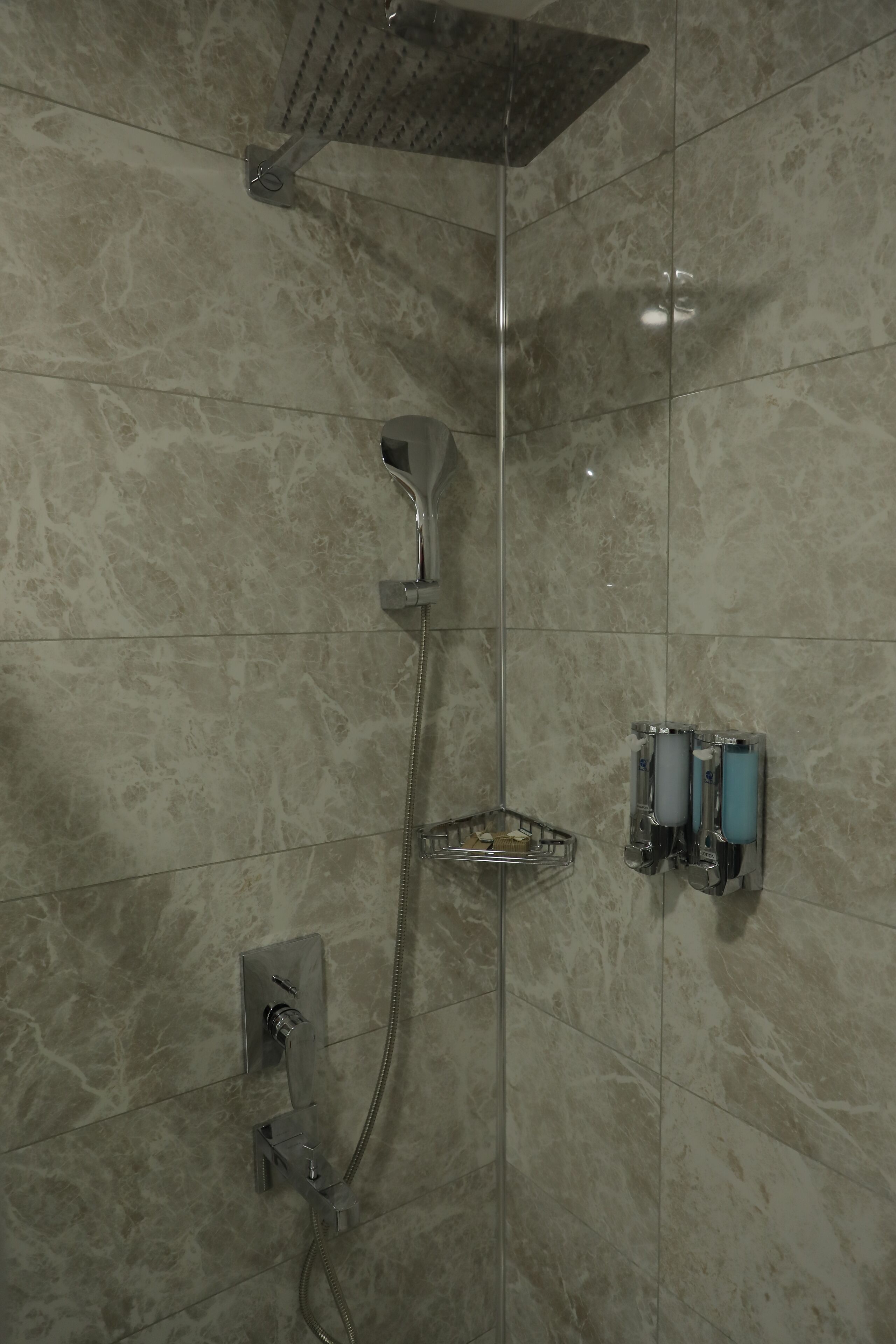 Área de chuveiro com chuveiro manual, torneira fixada na parede e saboneteira de vidro.