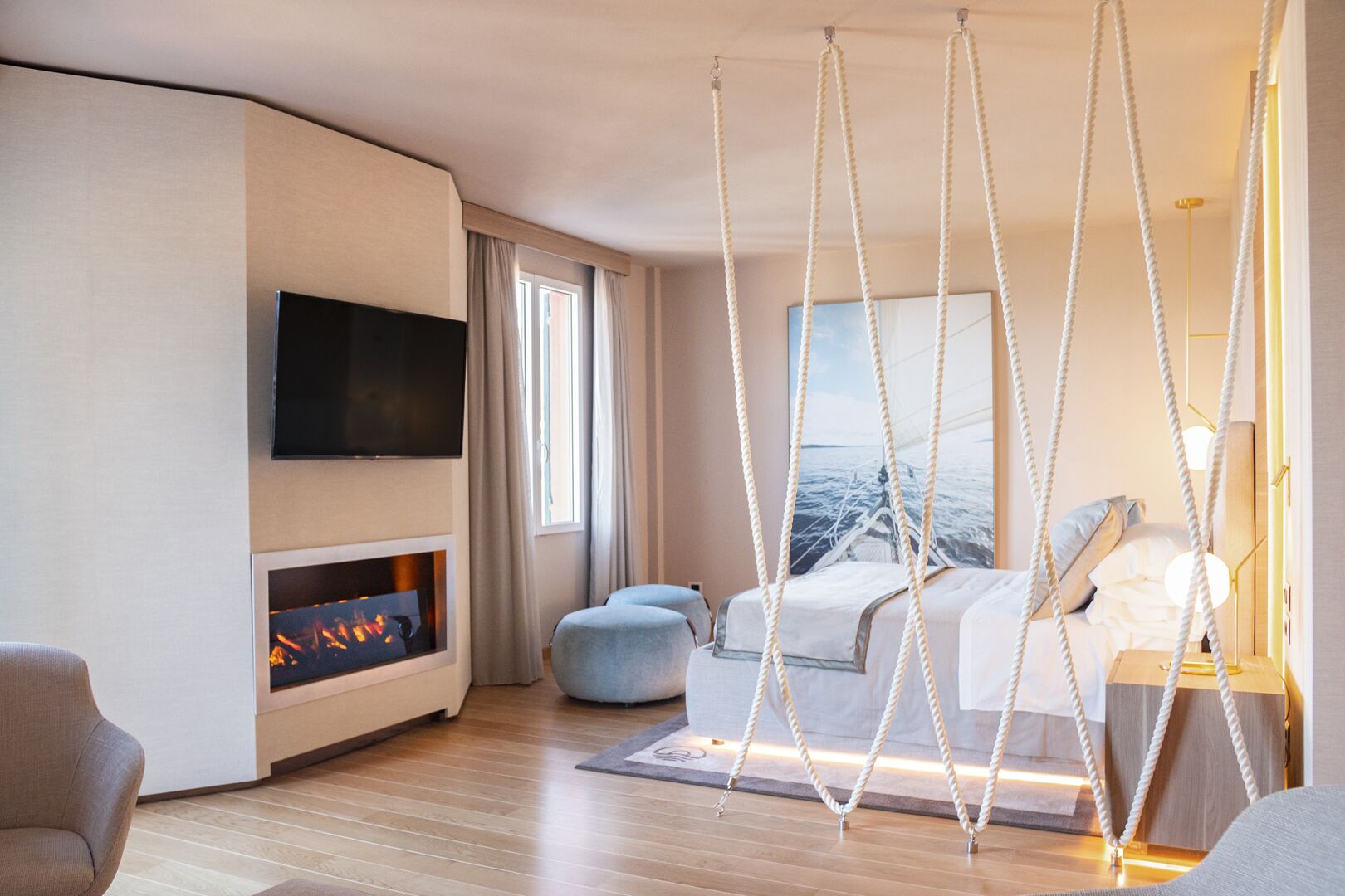 Quarto de hotel moderno com TV de tela plana, lareira, cama com vista para o mar e luminária pendente em corda.