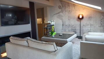 Uma sala de estar moderna com um sofá, uma mesa de centro e uma televisão. Há uma área de jantar com uma mesa e cadeiras. O quarto tem uma parede estampada e um abajur.