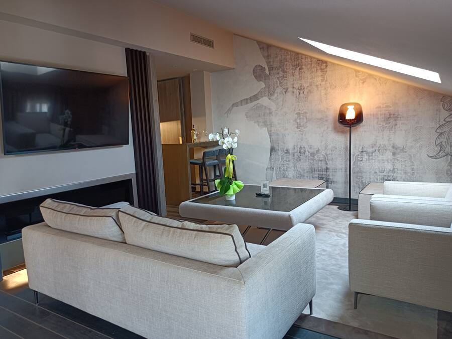 Uma sala de estar moderna com um sofá, uma mesa de centro e uma televisão. Há uma área de jantar com uma mesa e cadeiras. O quarto tem uma parede estampada e um abajur.