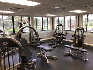 Academia com diversos equipamentos de ginástica, amplas janelas e vista para o verde lá fora.