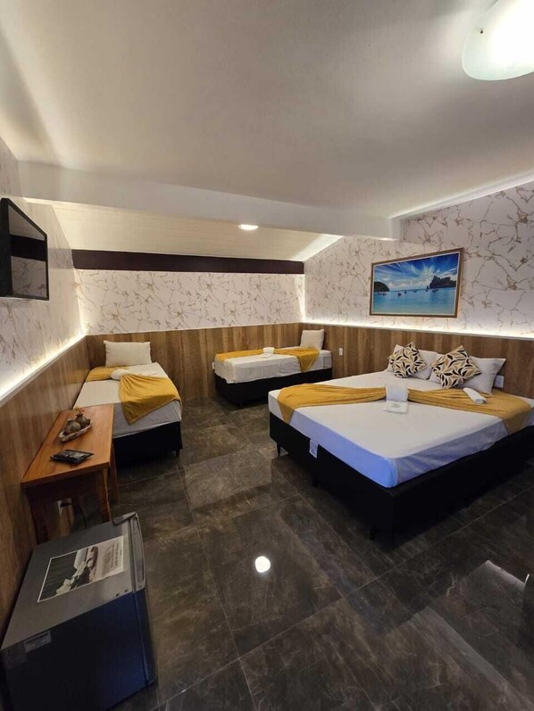 Quarto de hotel com três camas, uma escrivaninha, uma cadeira e uma pequena mesa com uma revista.