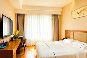 Quarto de hotel com cama, escrivaninha, cadeira, televisão e janela com cortinas.