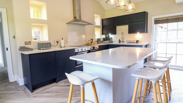 Uma cozinha moderna com ilha central, banquetas, diversos eletrodomésticos e iluminação pendente.