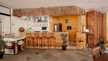 Uma área de bar com balcão, banquetas e geladeira. Há garrafas na parede e um extintor de incêndio.