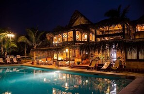 Resort de dois andares com piscina, espreguiçadeiras e uma estrutura com telhado de palha.