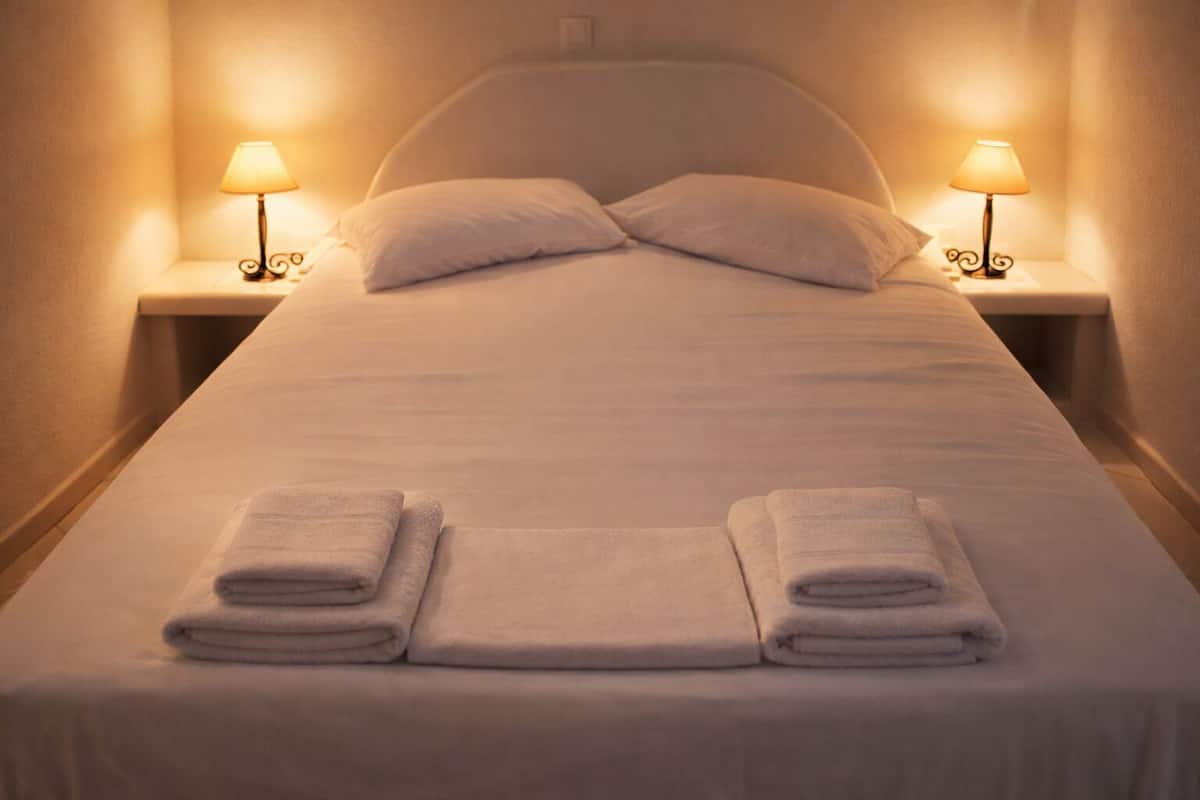 Cama bem arrumada com duas almofadas e duas toalhas dobradas no rodapé. Existem duas luminárias de cabeceira, uma de cada lado da cama.