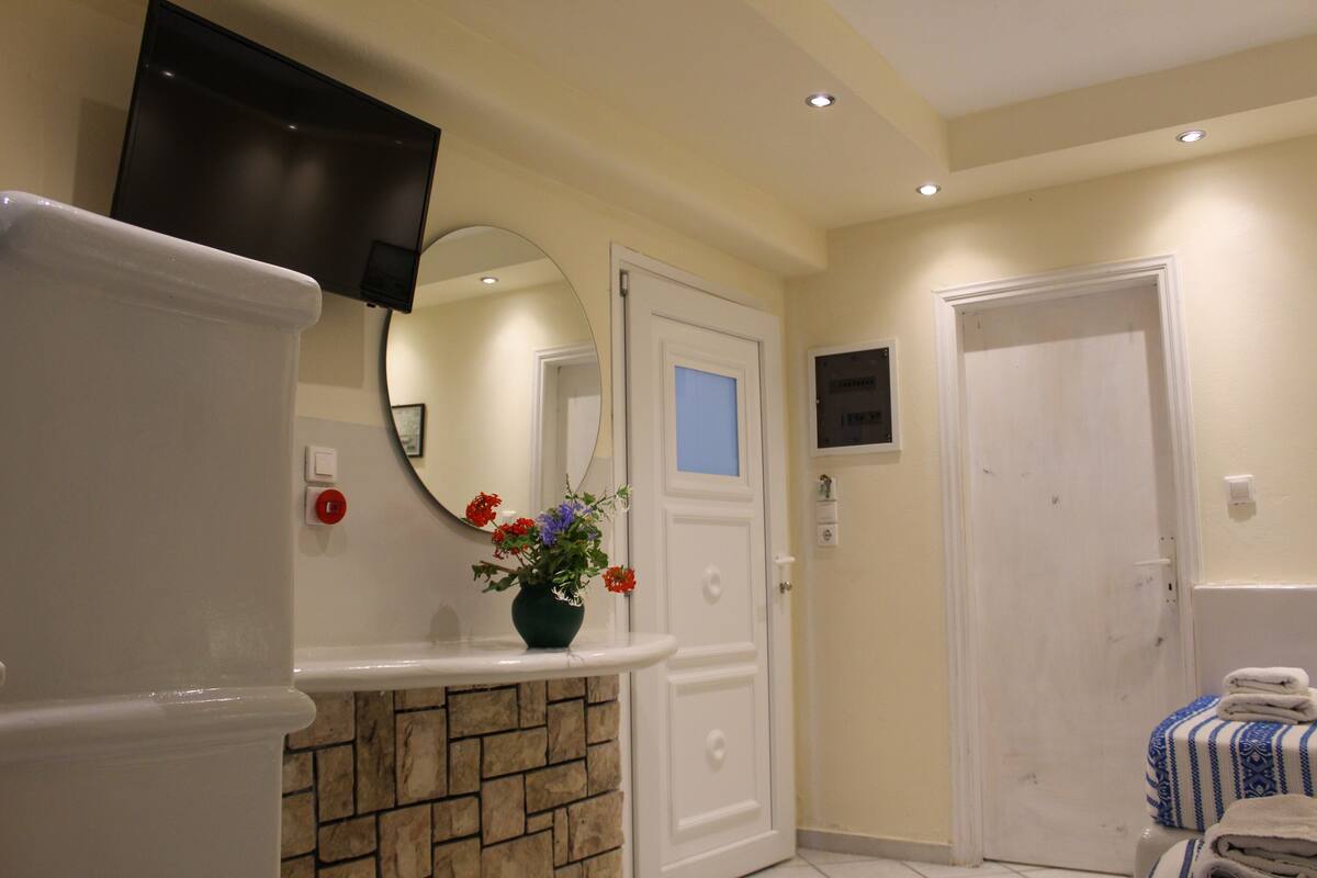 Casa de banho com toucador branco, espelho, televisão montada acima do espelho, porta com janela e um porta-toalhas pequeno com toalhas dobradas.