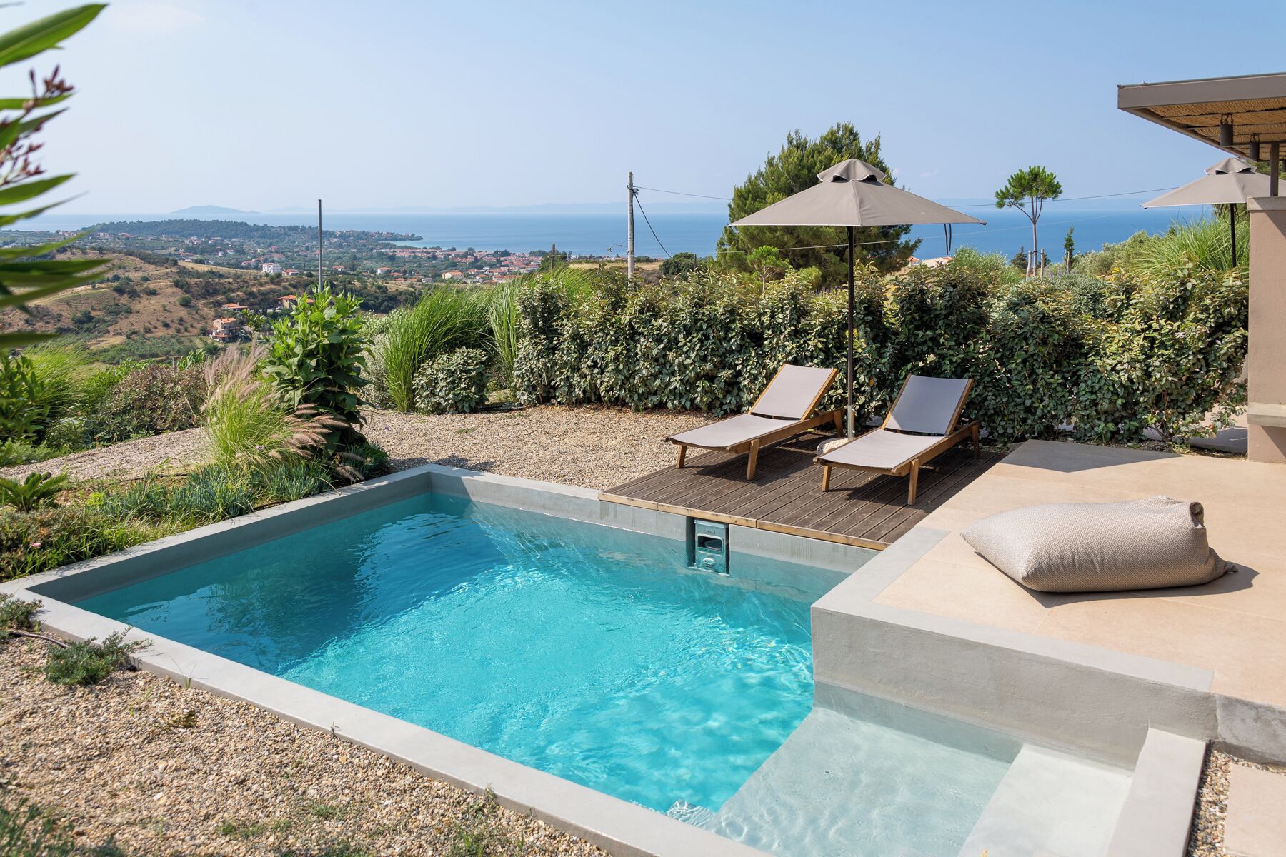 Uma piscina retangular com um deque de madeira, duas espreguiçadeiras e um guarda-sol coberto.