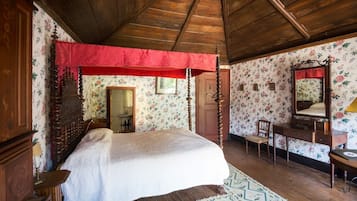 Um quarto com uma cama de dossel, teto de madeira, papel de parede floral, um espelho, cômoda e uma cadeira.
