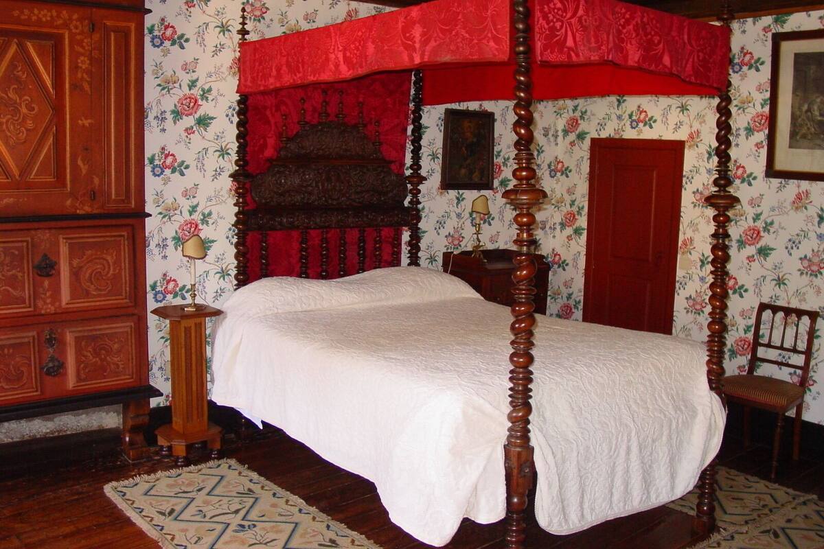 Uma cama com dossel e cortina vermelha, guarda-roupa de madeira, uma cadeira e papel de parede floral.