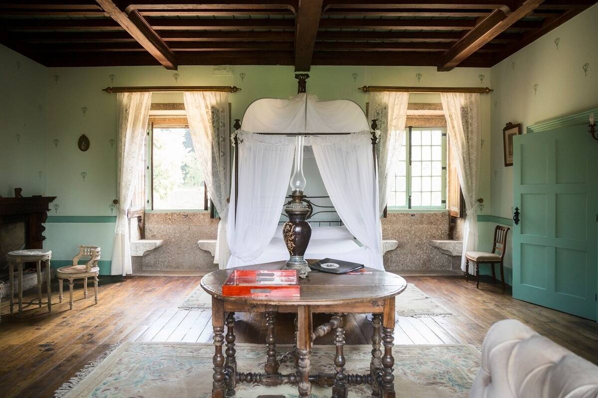 Um quarto com piso de madeira, uma mesa redonda com um objeto vermelho sobre ela, uma cadeira e uma cama com cortinas brancas.