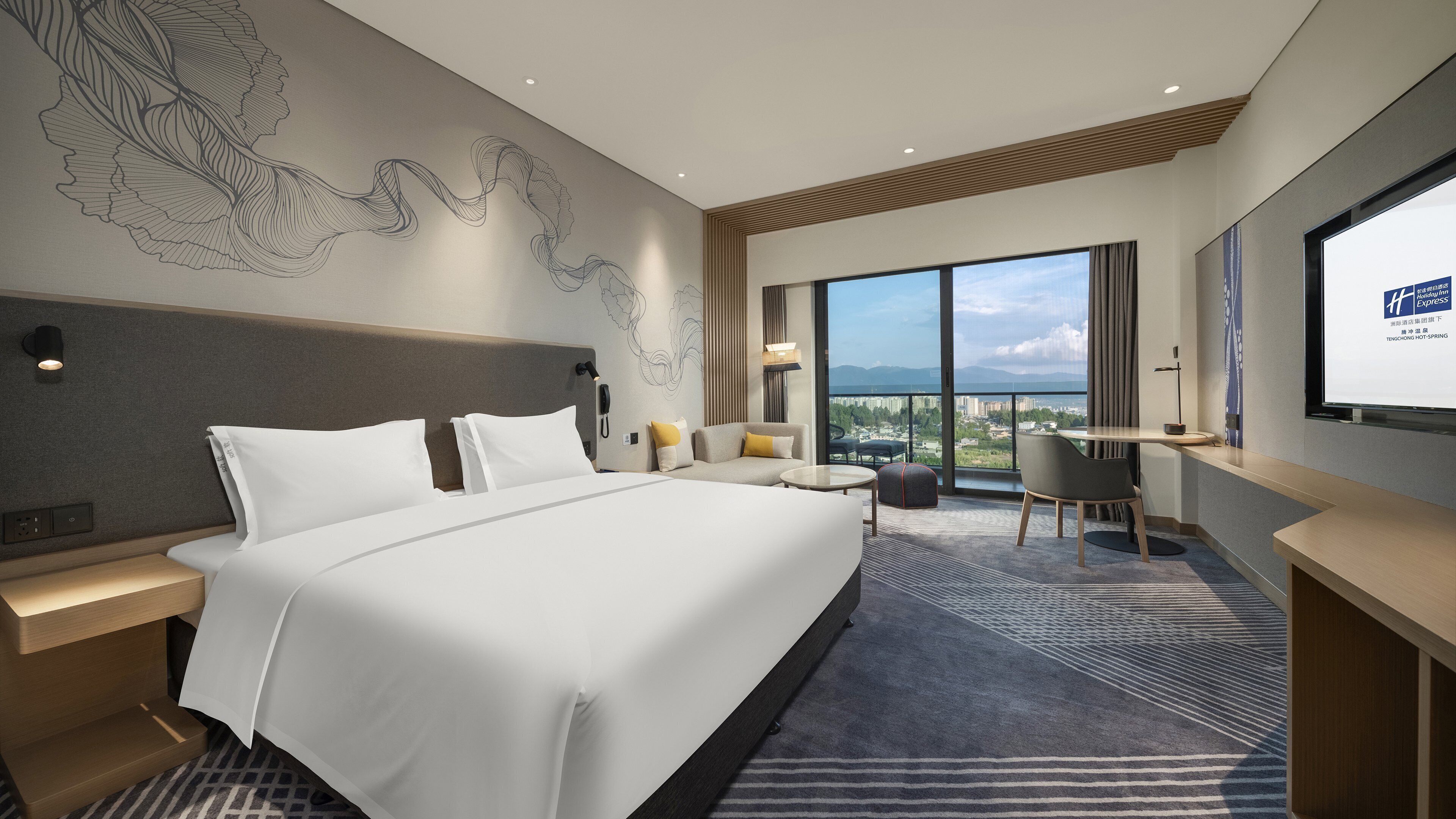 Quarto de hotel com uma cama grande, uma escrivaninha com cadeira, um sofá pequeno e uma janela ampla com vista para a cidade.