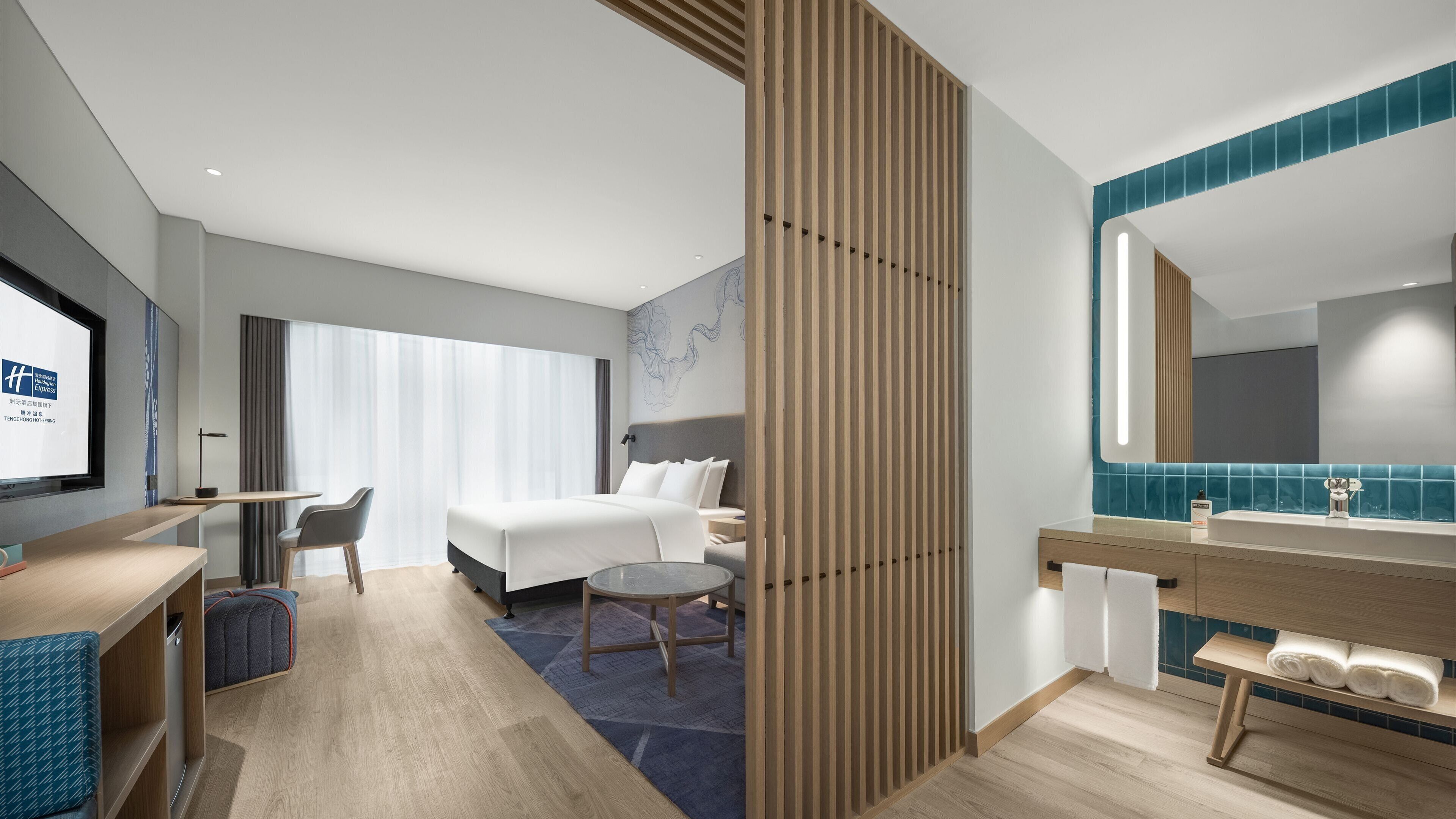 Quarto de hotel moderno com cama, mesa de cabeceira, cadeira, escrivaninha e banheiro com pia e espelho.