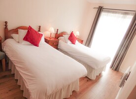 Quarto de hotel com duas camas de solteiro, cada uma com roupa de cama branca e almofadas vermelhas. Há mesas de cabeceira de madeira com luminárias, um cabeceira de madeira e uma janela com cortinas.