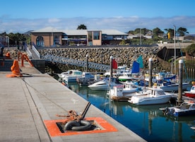Uma marina com barcos atracados, um muro de pedra e edifícios ao fundo.