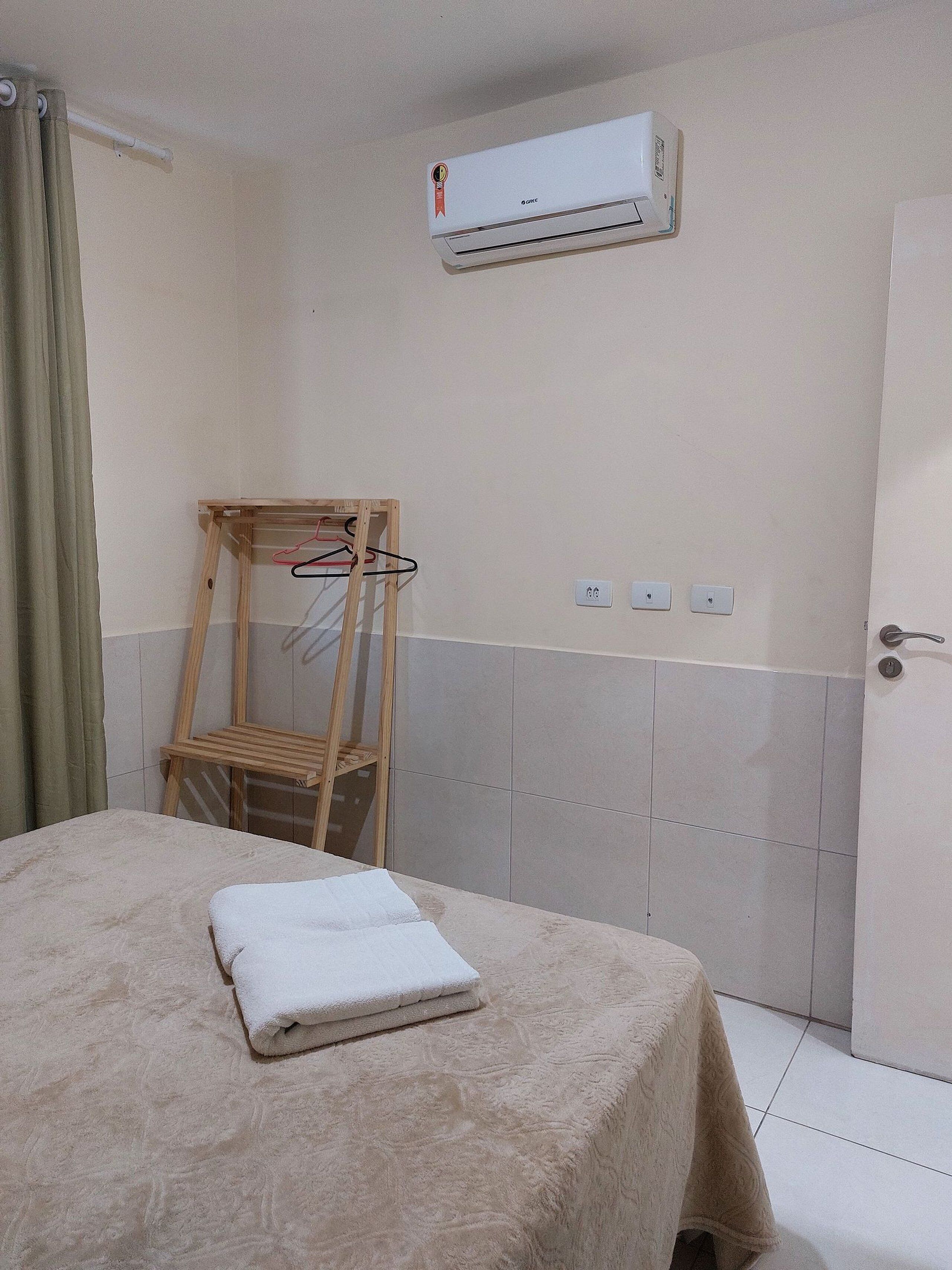Quarto de hotel com cama, ar condicionado, uma cadeira de madeira e toalha sobre a cama.