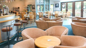 Um café moderno com mesas redondas e cadeiras estofadas, uma área de bar com banquetas e janelas amplas que permitem a entrada de luz natural.