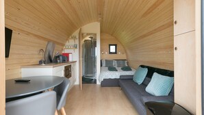 Um espaço interior compacto e de madeira com cozinha compacta, chuveiro, cama com roupa de cama verde e branca e sofá cinzento.