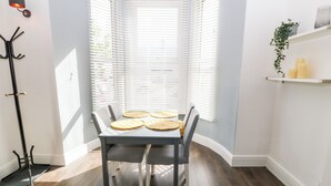 Uma sala de jantar com uma mesa posta para quatro pessoas, rodeada por cadeiras, com piso de madeira e paredes brancas.