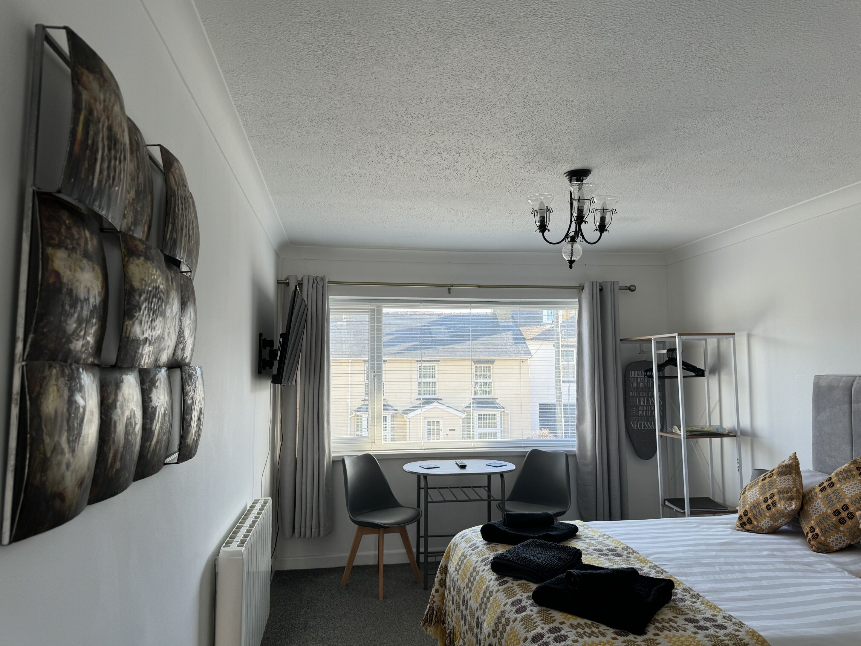 Quarto de hotel com cama, mesa, cadeira, radiador, janela com cortinas e um candelabro.