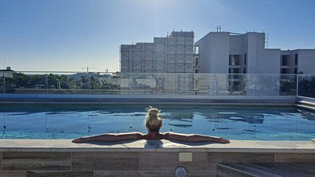 Uma pessoa relaxando à beira da piscina com vista para edifícios modernos e um céu claro.