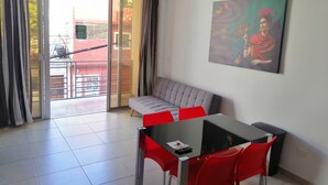 Quarto com porta de vidro deslizante para uma varanda, sofá cinza, mesa de vidro com cadeiras vermelhas e um quadro na parede.