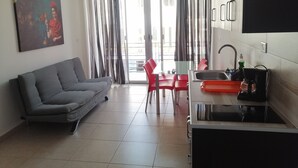 Uma sala moderna com um sofá cinza, uma área de jantar com cadeiras vermelhas, uma cozinha com pia e fogão, e uma janela grande com cortinas.