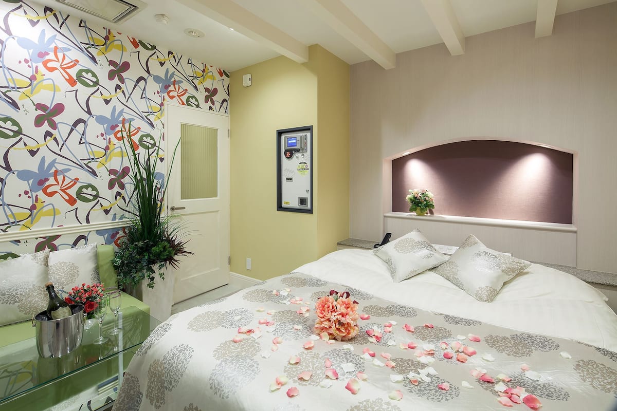 Quarto de hotel com uma cama, uma mesa de vidro com uma garrafa e flores, uma parede colorida e uma pequena planta.