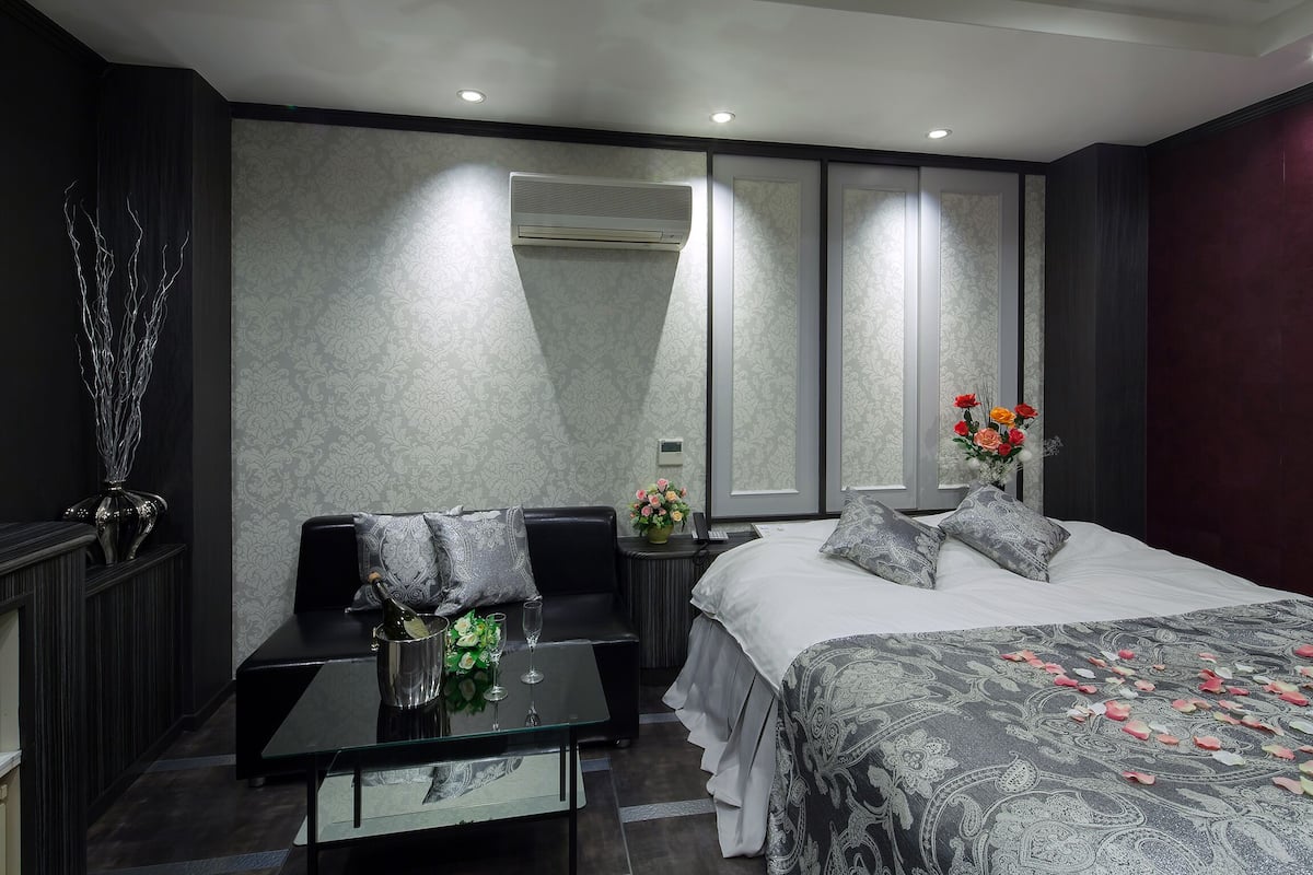 Quarto de hotel moderno com uma cama grande, um espaço de estar com mesa de vidro e uma mesa de cabeceira com um vaso de flores.