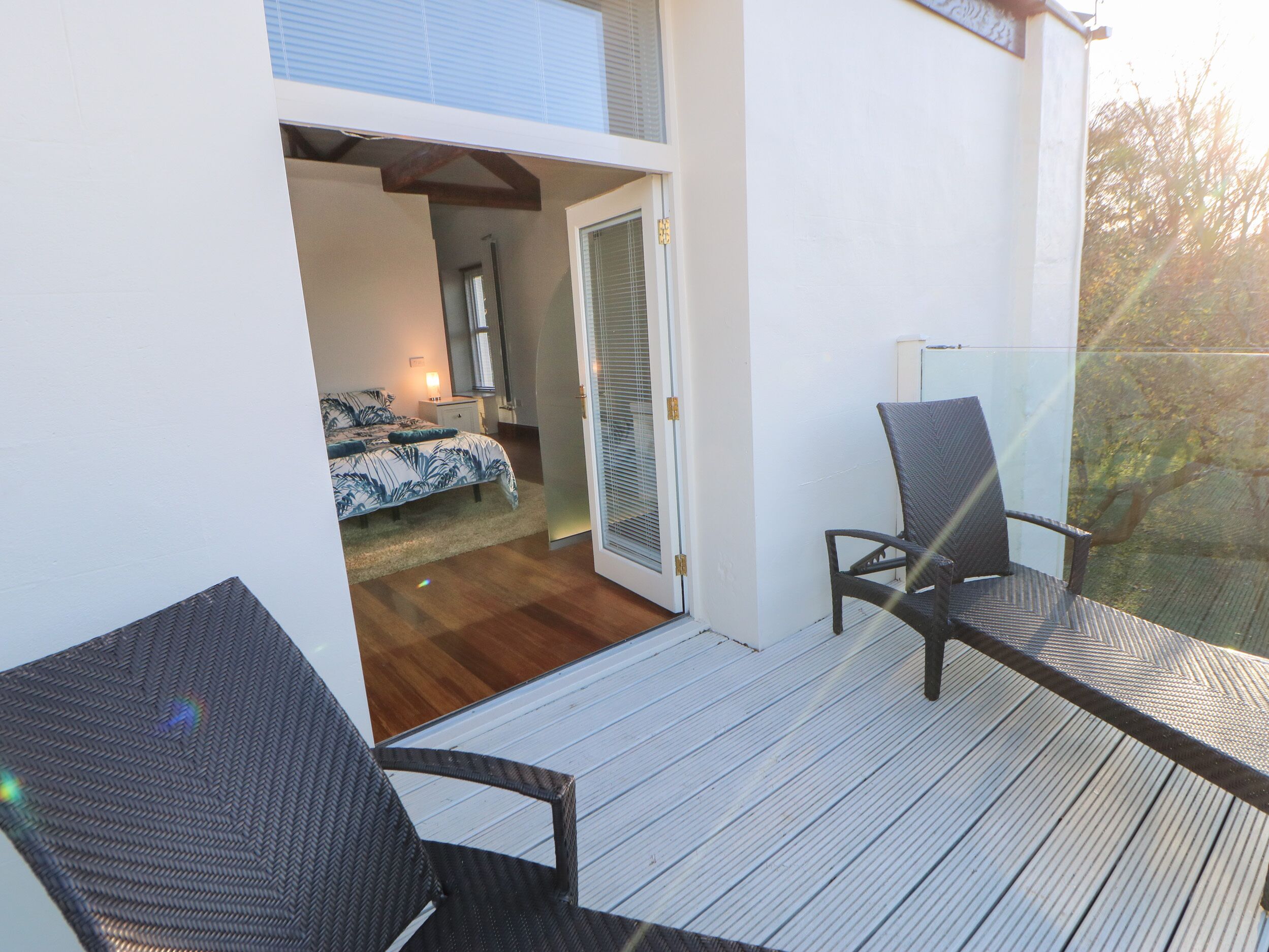Varanda com duas cadeiras de vime, um deck de madeira e um quarto com cama e mesa de cabeceira.
