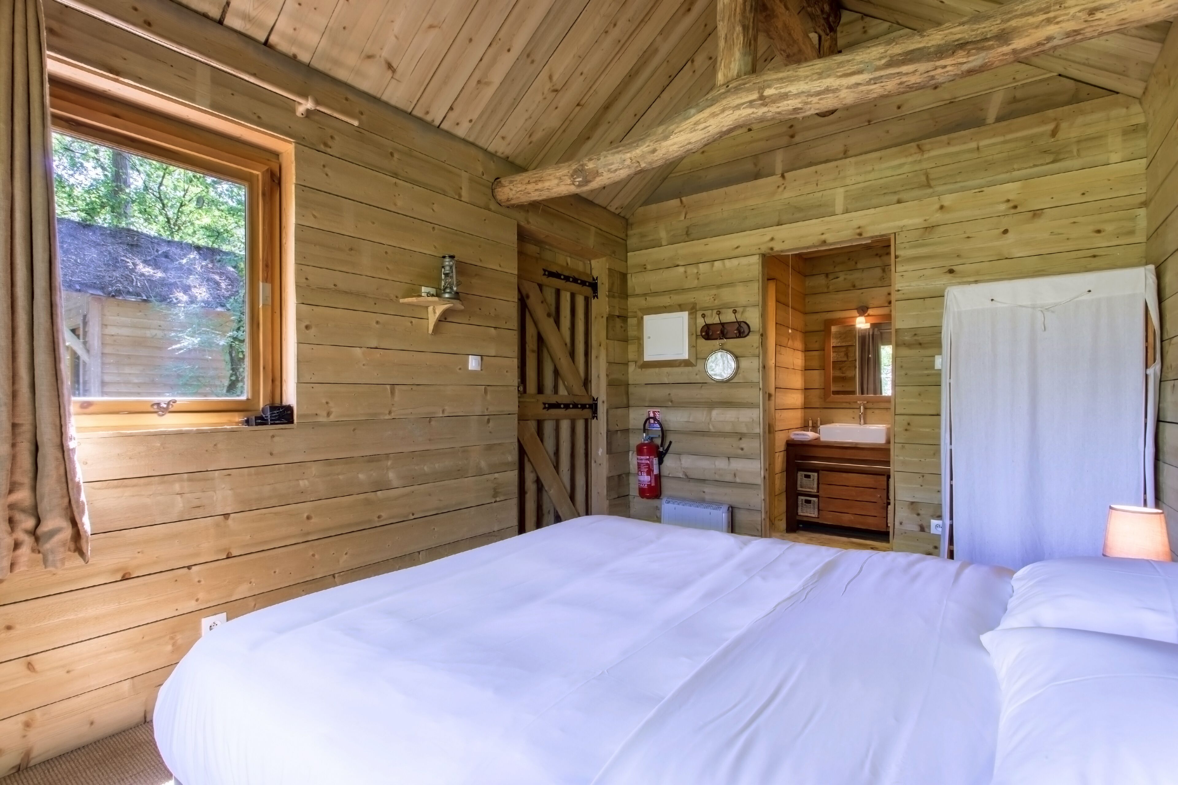 Quarto de cabana de madeira com cama, janela, espelho e banheiro com pia e vaso sanitário.