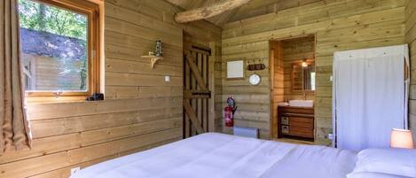 Quarto de cabana de madeira com cama, janela, espelho e banheiro com pia e vaso sanitário.