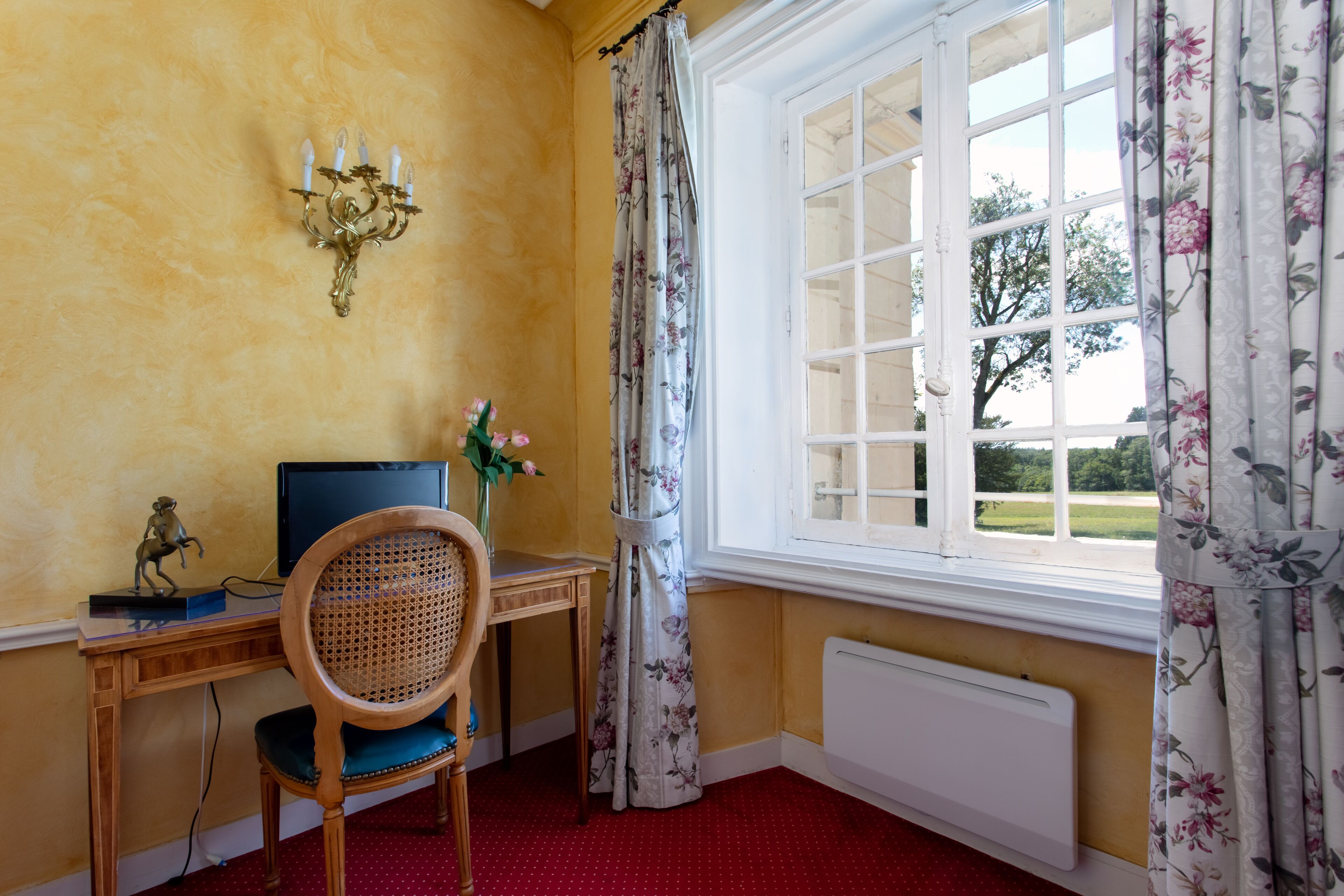 Quarto com escrivaninha de madeira, uma cadeira, televisão, radiador e janela com cortinas florais.