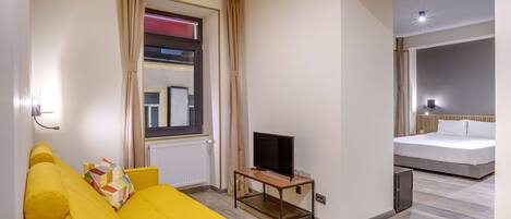 Quarto de hotel com um sofá amarelo, uma televisão sobre um suporte de madeira, uma cama e uma janela com cortinas.